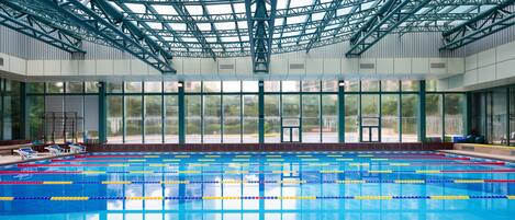 Piscina interior com pistas demarcadas, teto alto com estrutura metálica e grandes janelas com vista para o exterior.