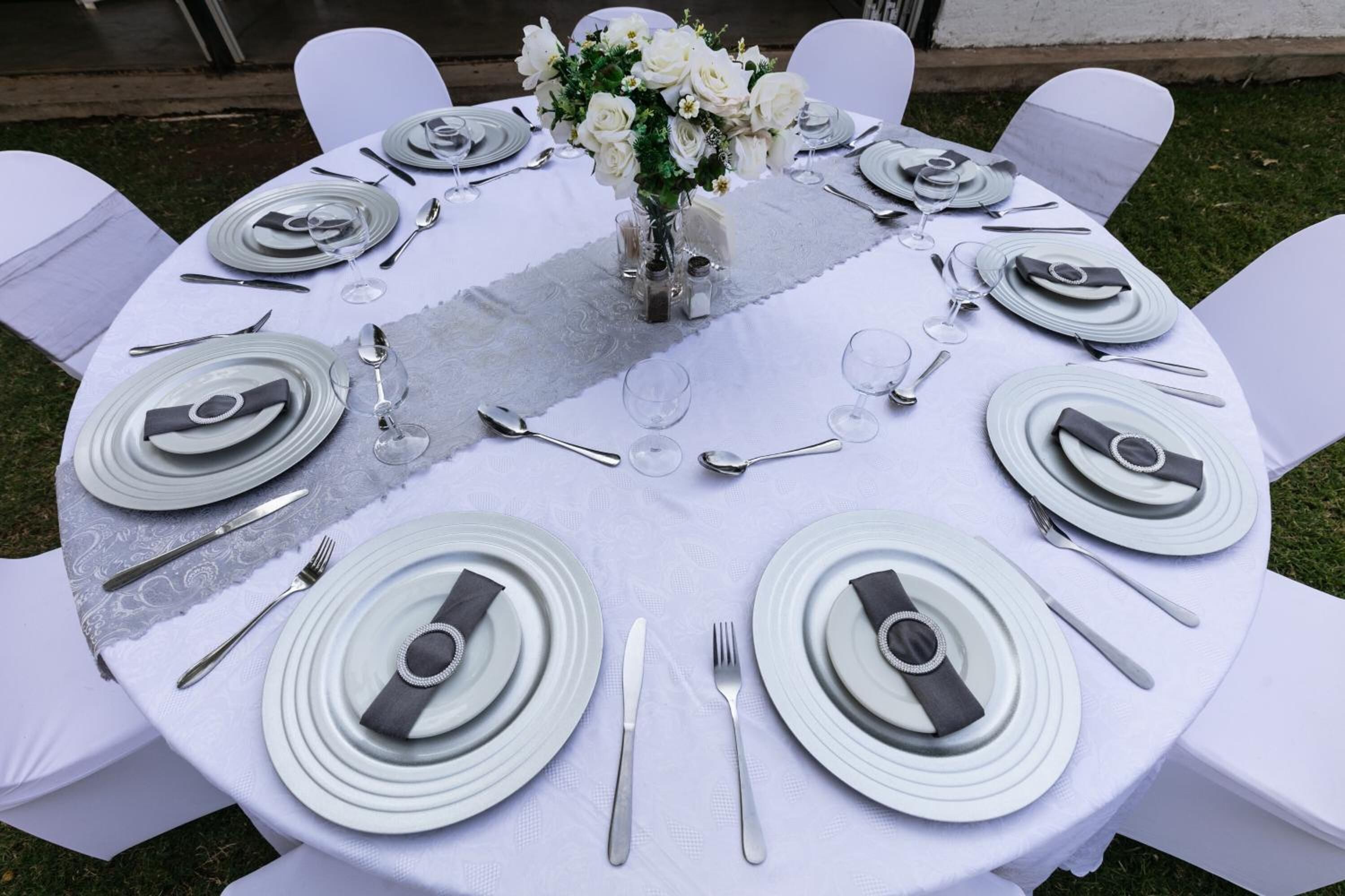Mesa redonda posta para um evento formal, com toalhas brancas, pratos, talheres e um vaso com flores brancas.