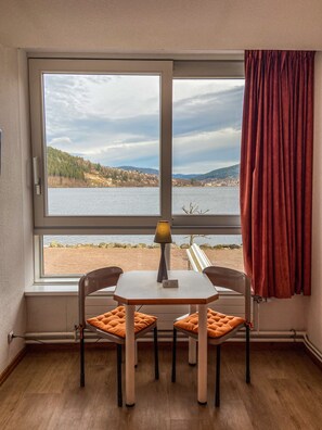 Quarto com uma janela grande que oferece vista para um lago e colinas ao longe. Há uma mesa com duas cadeiras e um abajur.