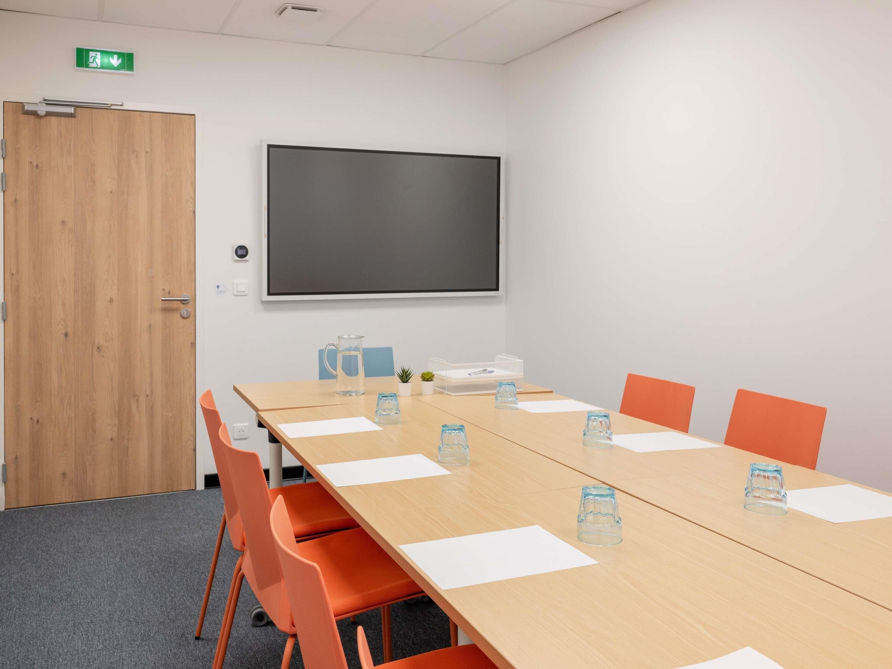 Uma sala de conferências com uma mesa comprida, cadeiras laranja, uma tela de projetor e uma porta com sinal de saída de emergência.