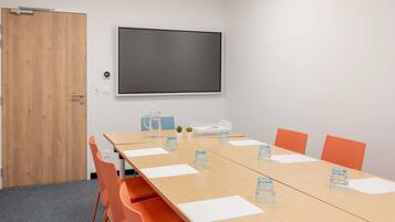 Uma sala de conferências com uma mesa comprida, cadeiras laranja, uma tela de projetor e uma porta com sinal de saída de emergência.