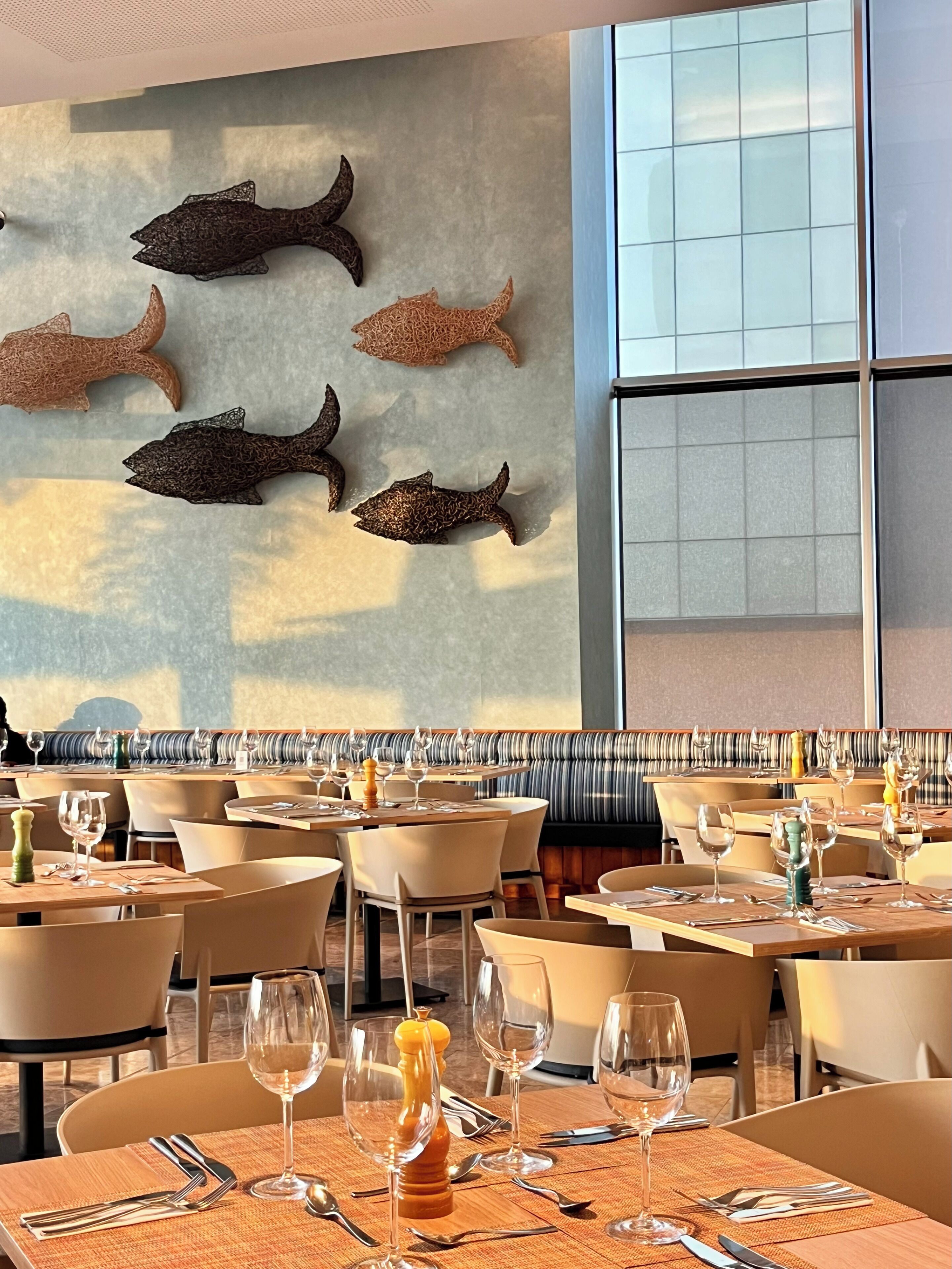 Interior de restaurante com mesas preparadas para refeições, uma grande janela e arte decorativa de peixes na parede.