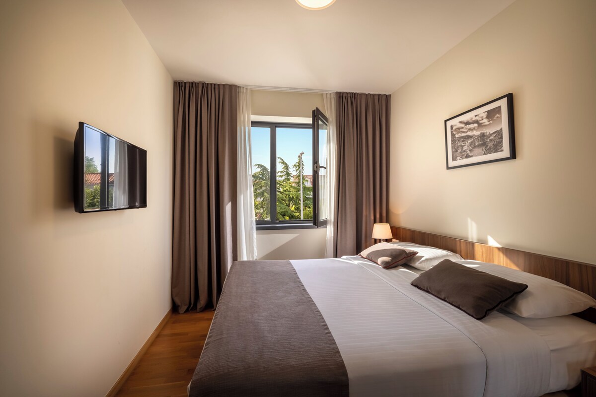 Quarto de hotel com cama, televisão na parede, janela com cortinas e um quadro na parede.