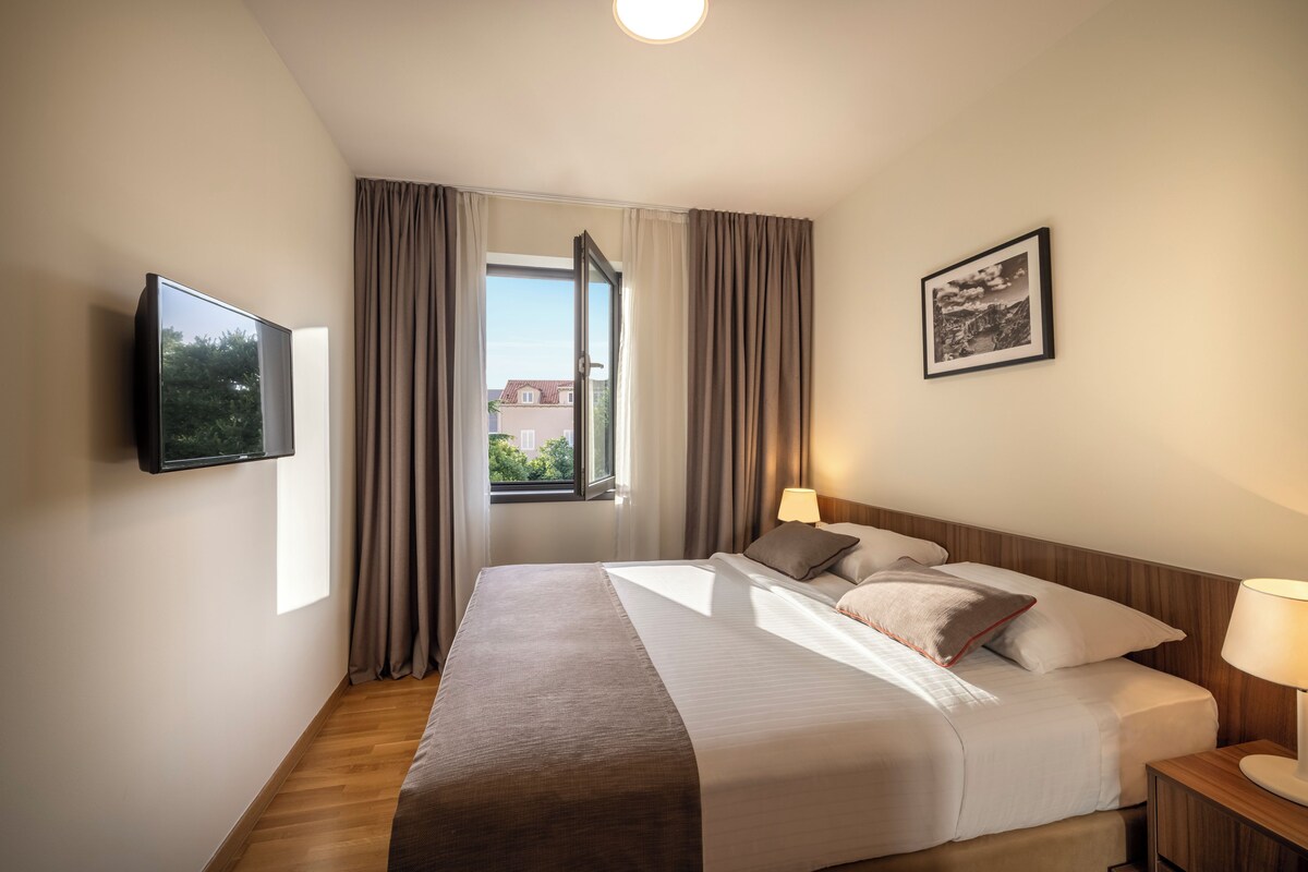 Quarto de hotel com cama, televisão, janela com cortinas, quadro na parede e mesa de cabeceira com abajur.