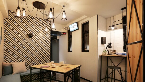 Quarto de hotel moderno com parede estampada, escrivaninha de madeira, um banco de bar e um espaço de estar com almofadas.