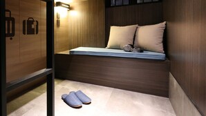 Quarto compacto com cama, almofadas, mesa de cabeceira e chinelos no chão.