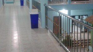 Um corredor comprido e estreito, com chão de azulejos, paredes azuis e uma grade de metal de um lado. Há lixeiras e uma escada que leva a um andar superior.