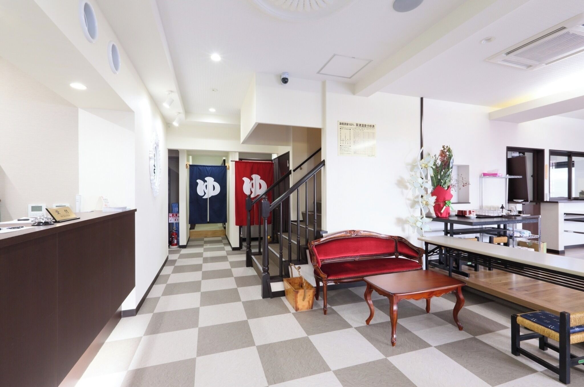 Um saguão de hotel com balcão de recepção, escada, sofá vermelho, mesa de madeira e uma longa mesa de jantar.