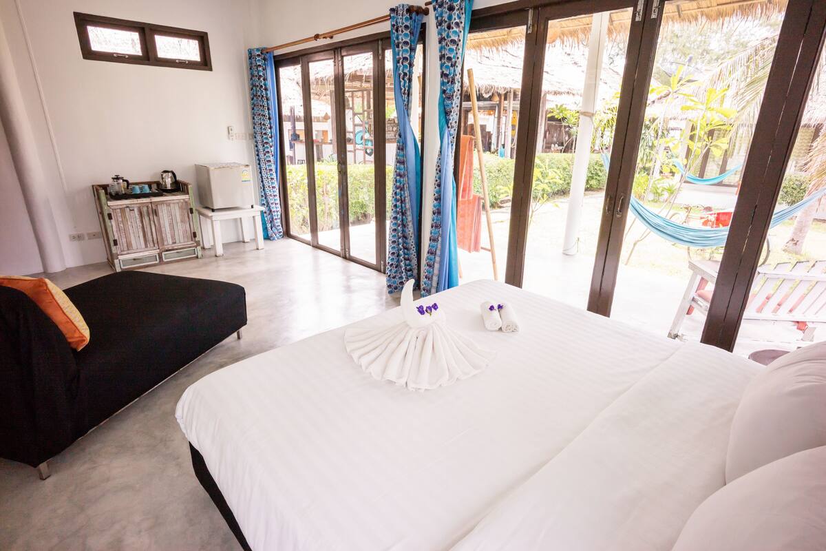 Quarto de hotel com uma cama grande, um sofá pequeno, um minibar e vista para a área externa através de amplas portas de vidro deslizantes.