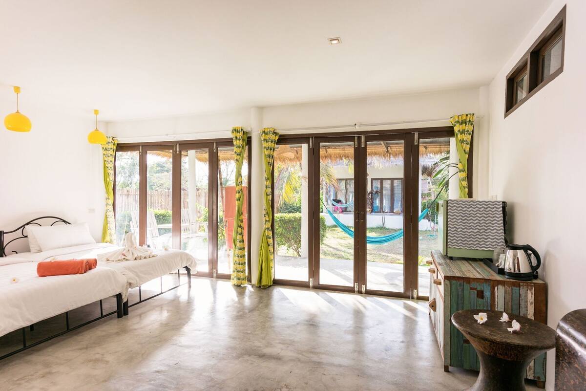 Quarto com cama, uma mesinha com chaleira e vista para um jardim através de grandes portas de vidro.
