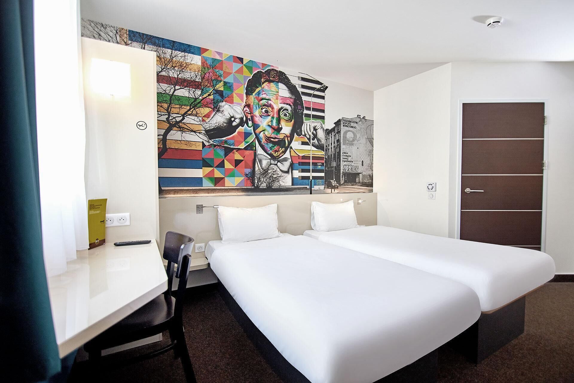 Quarto de hotel com uma cama grande, uma escrivaninha com cadeira, televisão na parede e uma porta.