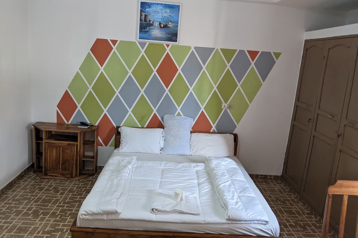 Cama bem arrumada com lençóis brancos, cabeceira de madeira e parede atrás com um padrão geométrico em losangos coloridos.
