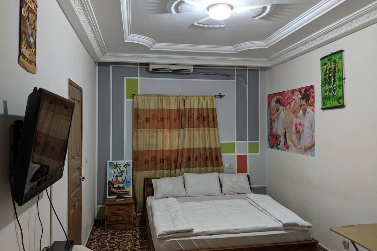 Quarto com cama, televisão sobre suporte, pintura na parede e ventilador de teto.