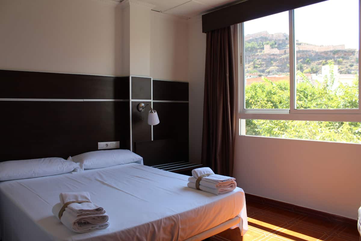 Quarto de hotel com uma cama, mesa de cabeceira, luminária e janela com vista para vegetação e uma construção ao longe.