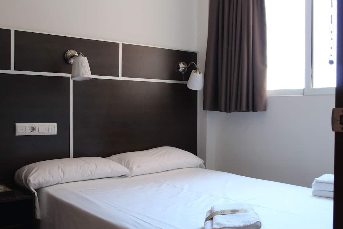Quarto de hotel com cama, duas luminárias na parede, janela com cortinas e mesa de cabeceira com abajur.