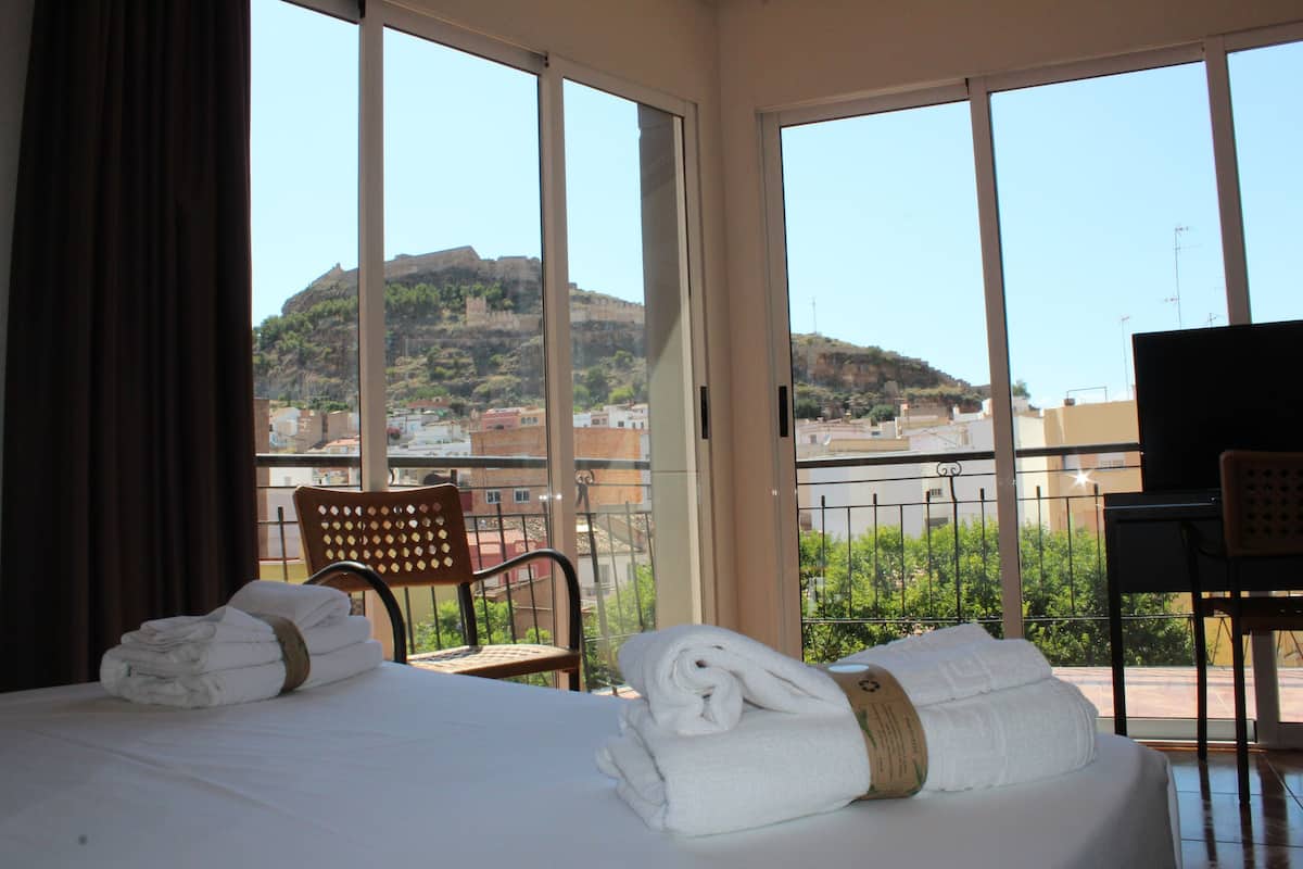 Quarto de hotel com cama, poltrona, escrivaninha e vista para uma colina.