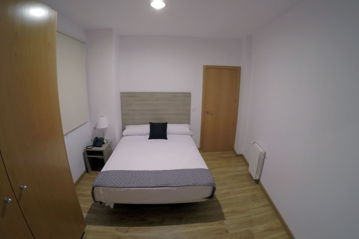 Quarto com cama, mesa de cabeceira com luminária, guarda-roupa de madeira e uma porta.