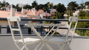Mesa e cadeiras brancas ao ar livre numa varanda, com vista para edifícios residenciais e vegetação.