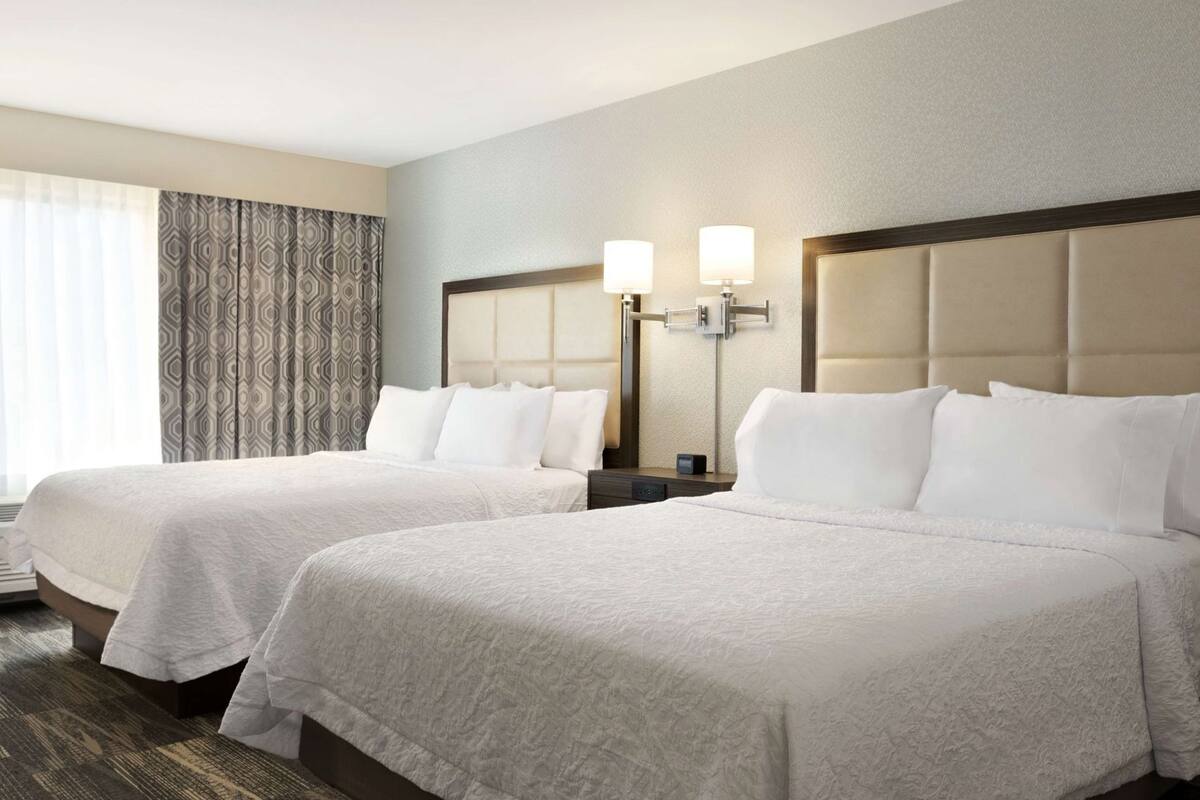 Quarto de hotel com duas camas, cabeceira com luminária e cortina estampada.