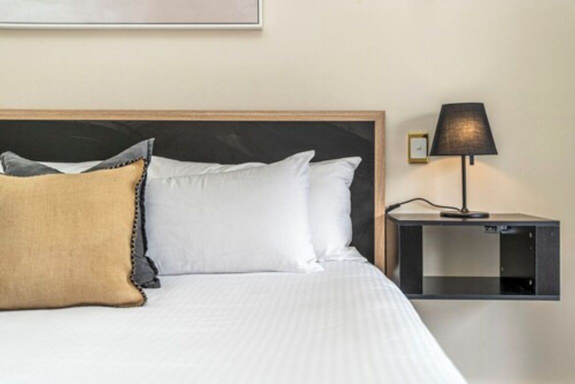 Cama bem arrumada com lençóis brancos e duas almofadas, mesa de cabeceira com abajur e quadro na parede.