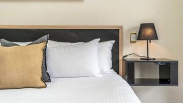 Cama bem arrumada com lençóis brancos e duas almofadas, mesa de cabeceira com abajur e quadro na parede.