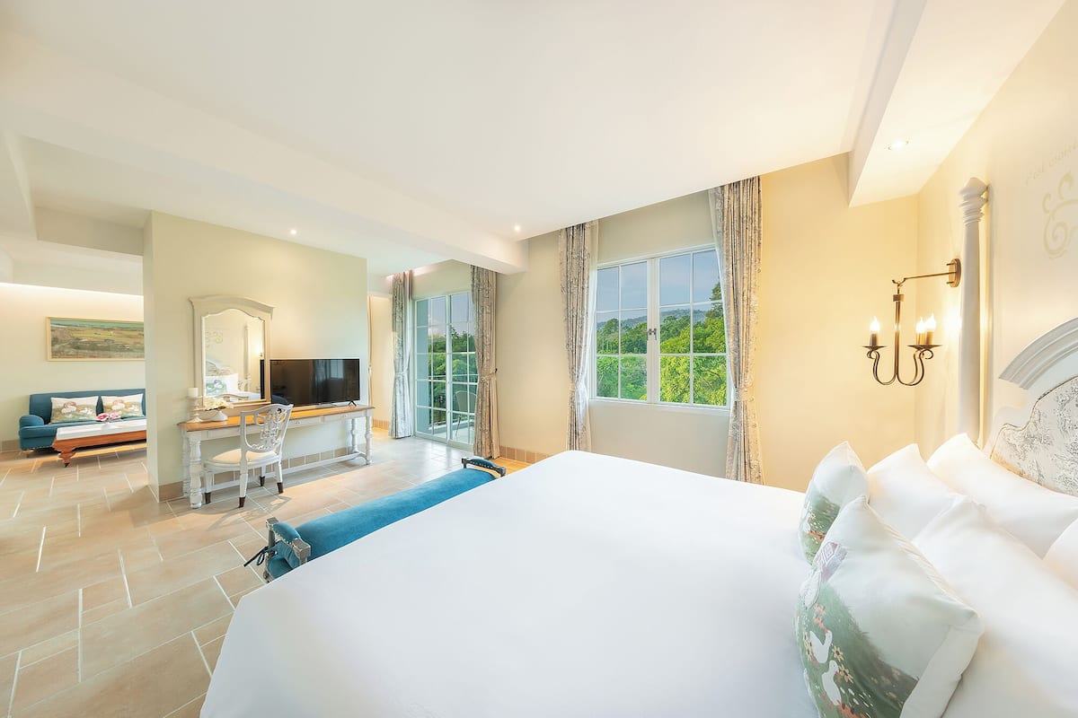 Quarto de hotel com uma cama grande, uma escrivaninha com TV, uma cadeira e vista para o verde lá fora.