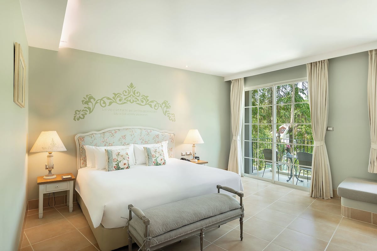 Quarto com cama, duas mesas de cabeceira, um banco, janela com cortinas e um objeto decorativo na parede.