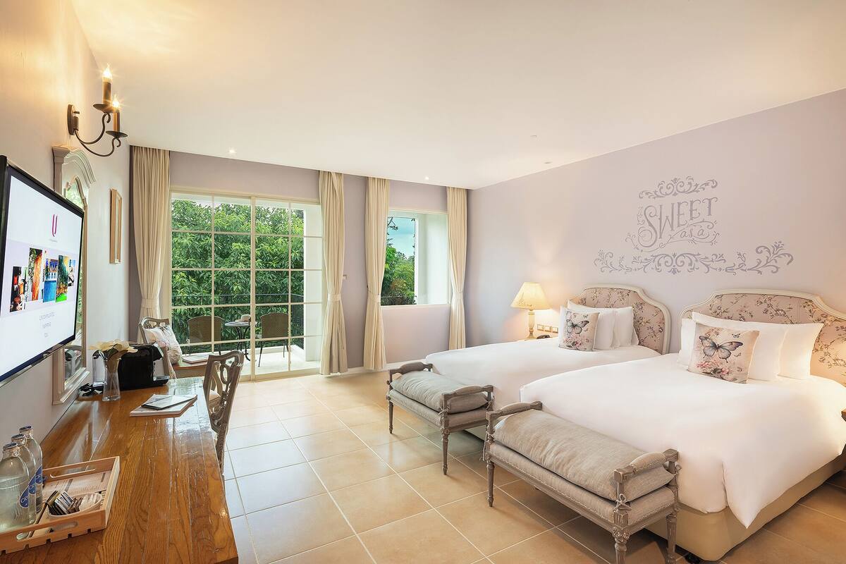Quarto de hotel com duas camas, uma escrivaninha com computador, uma janela com cortinas e uma parede com uma frase decorativa.