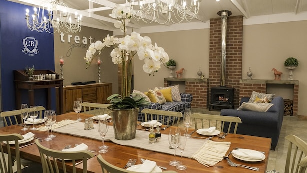 Uma sala de jantar com mesa de madeira posta para seis pessoas, cercada por cadeiras. Há uma lareira de tijolos, um sofá azul e um candelabro sobre a mesa.