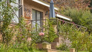 Uma casa de tijolos com varanda, janela com cortina e jardim com diversas plantas e flores.