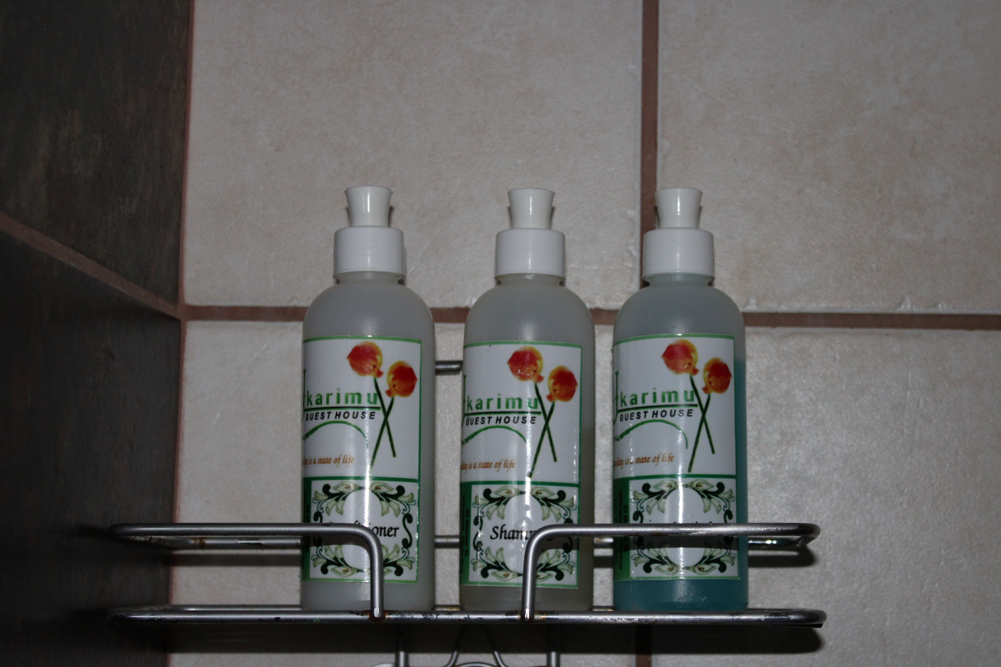 Três garrafas de shampoo da residência Karimyu sobre uma prateleira de metal em frente a uma parede revestida de azulejos.