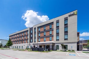 Um hotel moderno com uma placa que diz "HOME 2". Apresenta uma construção em vários andares com fachadas de tijolo e pedra, janelas amplas e uma área externa coberta com assentos.