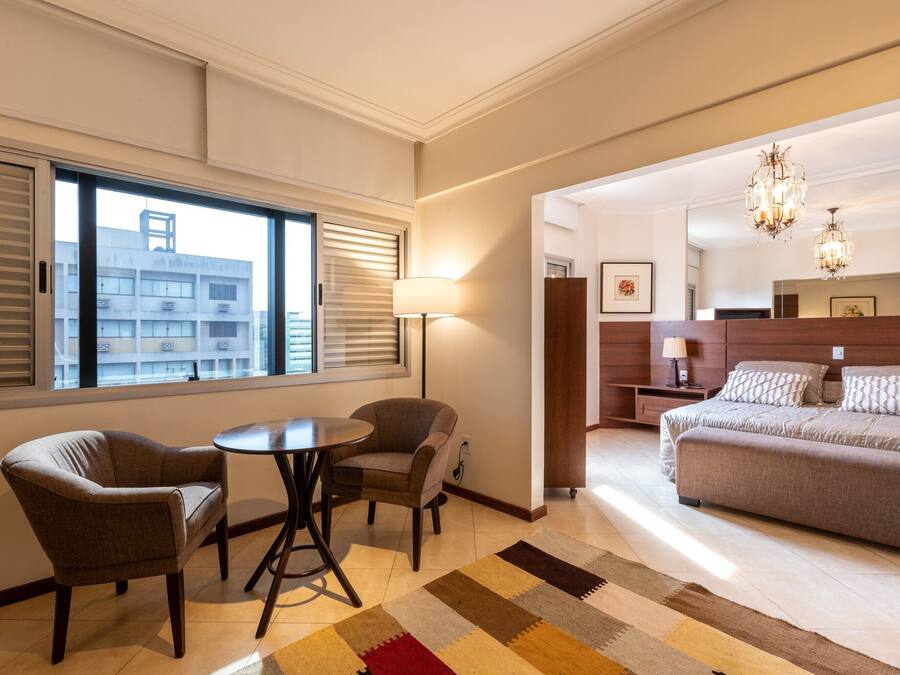Quarto de hotel com uma cama, duas poltronas, uma mesinha, uma luminária e vista para prédios pela janela.