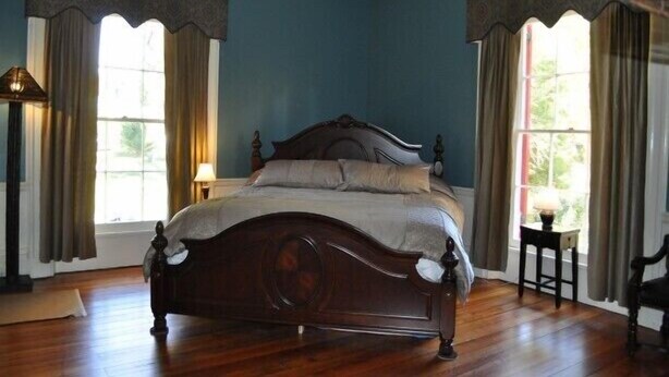 Quarto com uma cama grande, duas janelas com cortinas, uma mesa de cabeceira e um abajur.