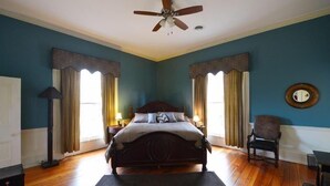 Quarto com cama de madeira, duas mesas de cabeceira, uma cadeira, ventoinha no teto e uma janela grande com cortinas.
