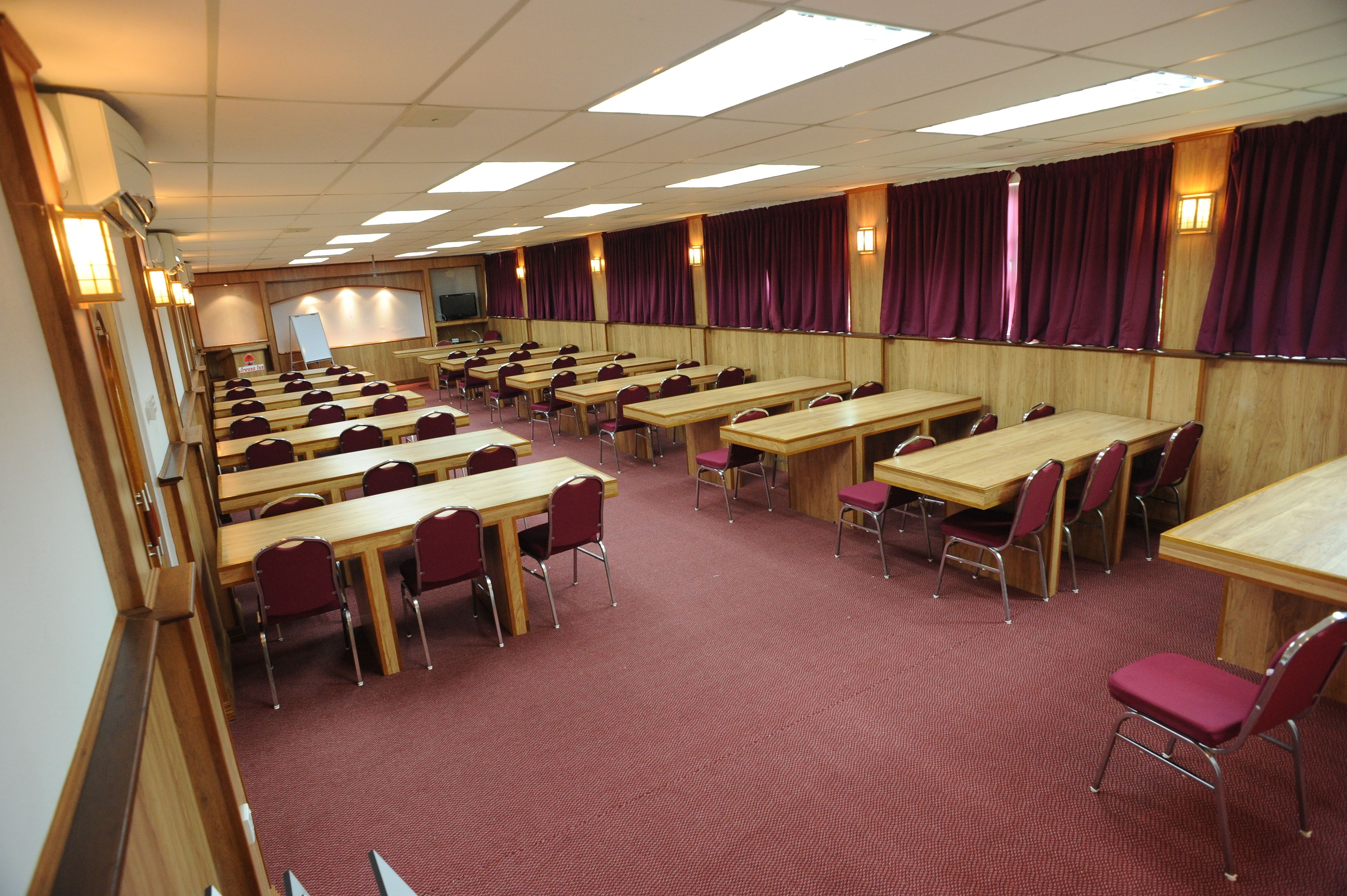 Sala de conferências com fileiras de mesas de madeira e cadeiras vermelhas, equipada com iluminação no teto e uma tela de projeção.