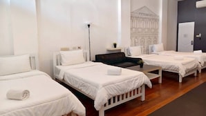 Quarto de hotel com três camas de solteiro, um sofá escuro, uma parede branca com um grande desenho arquitetônico e piso de madeira.