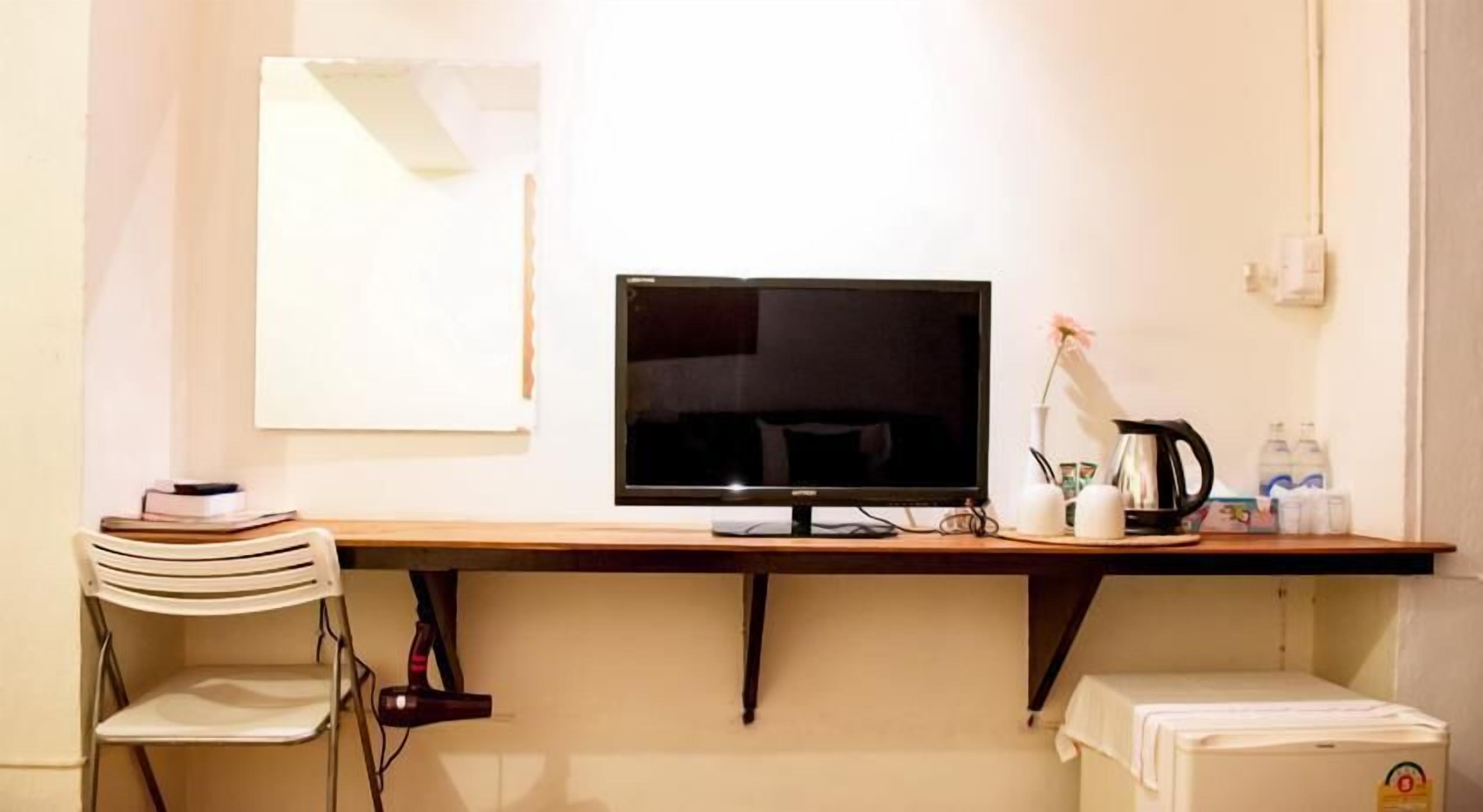 Quarto pequeno e bem iluminado, com escrivaninha, cadeira, televisão e um pequeno espaço de cozinha.