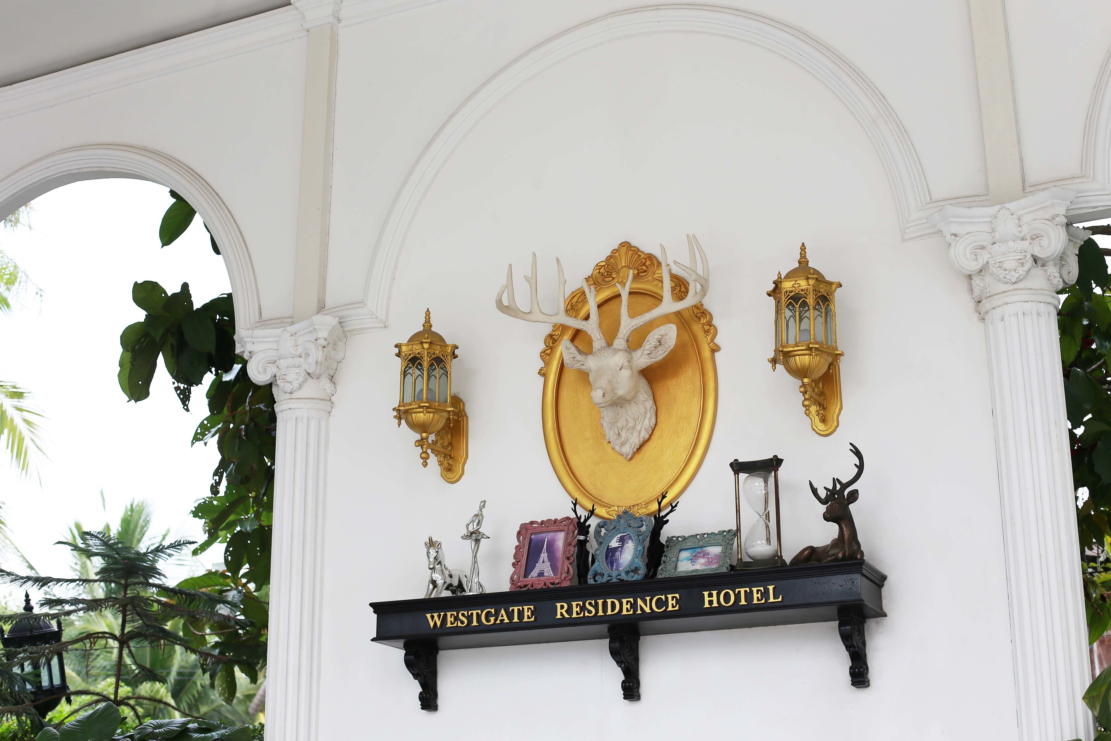Um letreiro fixado na parede com uma cabeça de veado e chifres, duas lanternas e uma estante com objetos emoldurados e um letreiro com a inscrição 'Westgate Residence Hotel'.