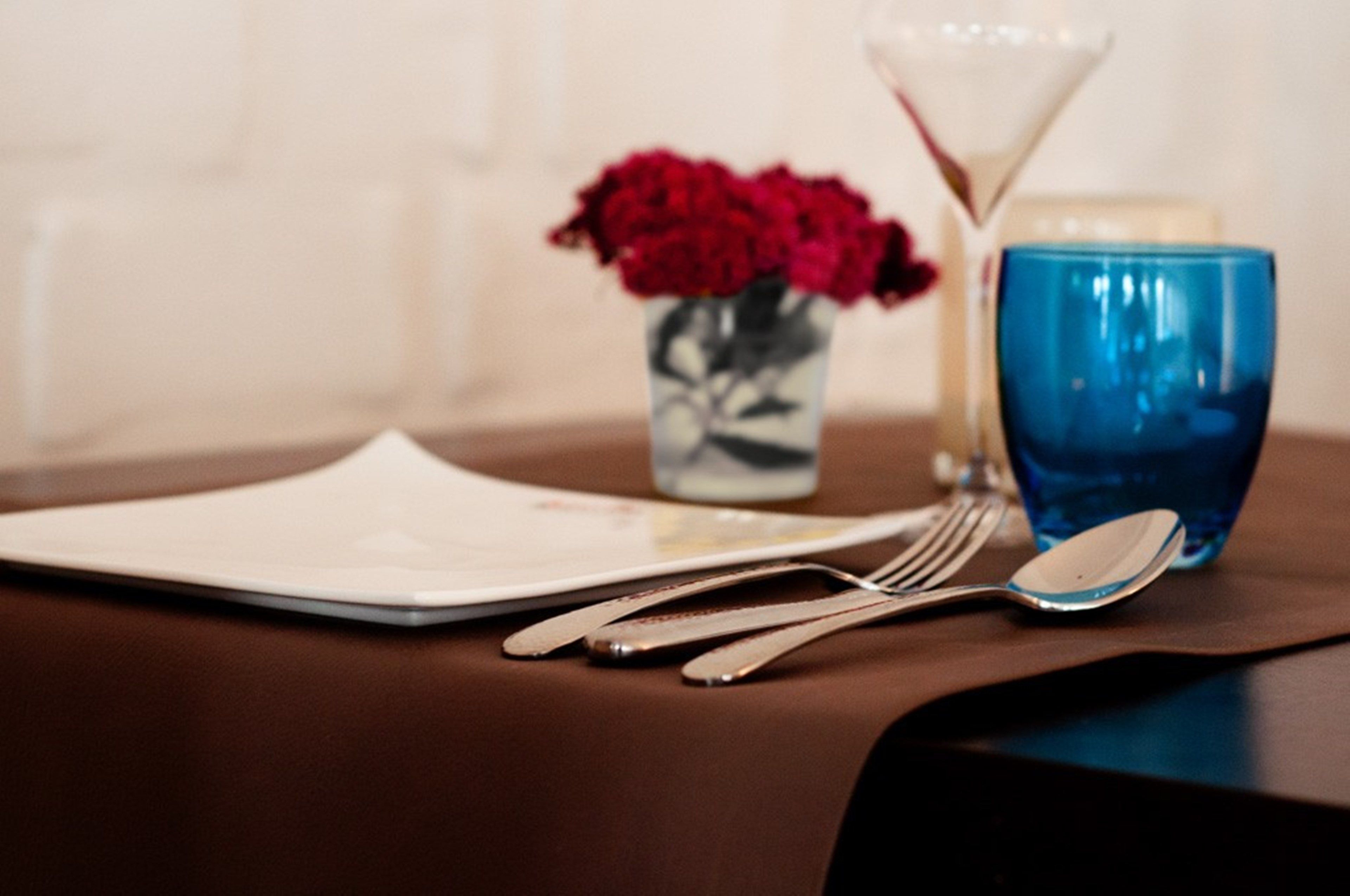 Uma mesa de jantar bem posta, com um prato branco, talheres de prata, um copo azul e um vaso com flores vermelhas.