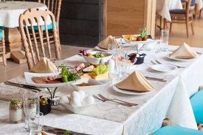 Uma mesa posta para o pequeno-almoço, com diversos pratos, incluindo ovos, frutas e pão, sobre uma toalha branca.