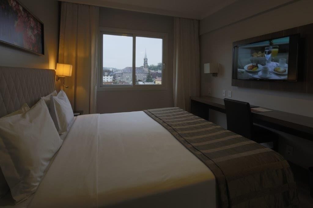 Quarto de hotel com cama, televisão, escrivaninha e janela com vista para a cidade.