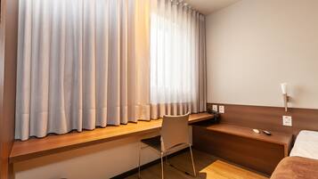 Quarto de hotel com escrivaninha de madeira, uma cadeira, uma cama e janela com cortinas.