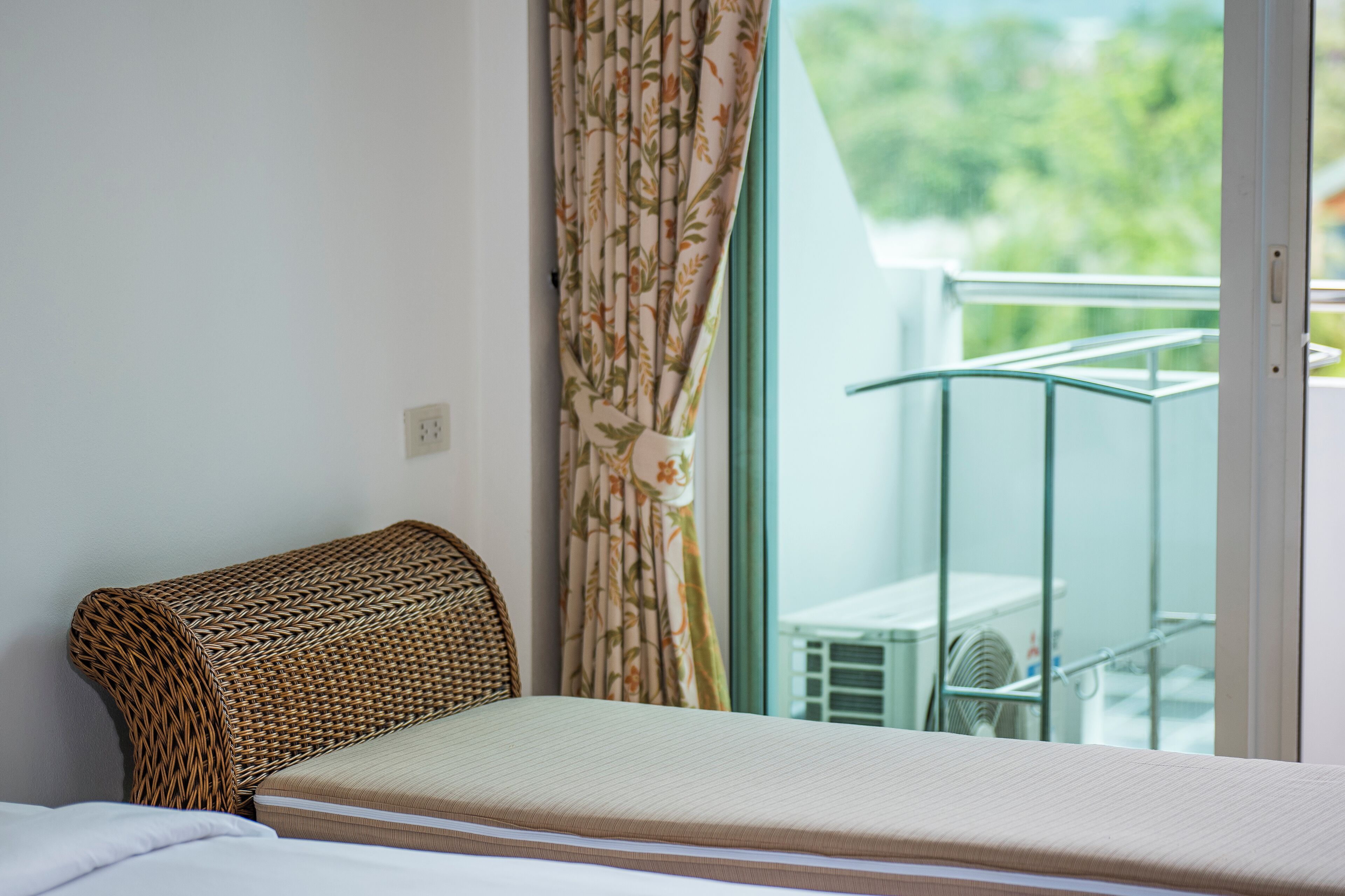 Um quarto com cama, uma cadeira de vime, uma janela com cortina e vista para uma varanda com grades.