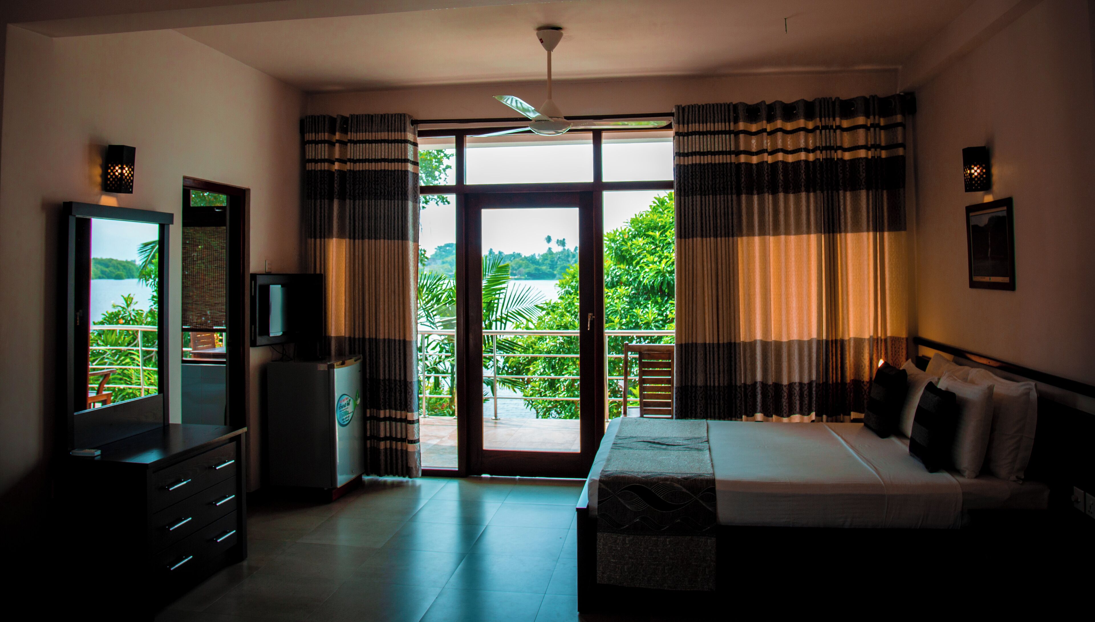 Quarto de hotel com uma cama, televisão, cómoda e varanda com cortinas.