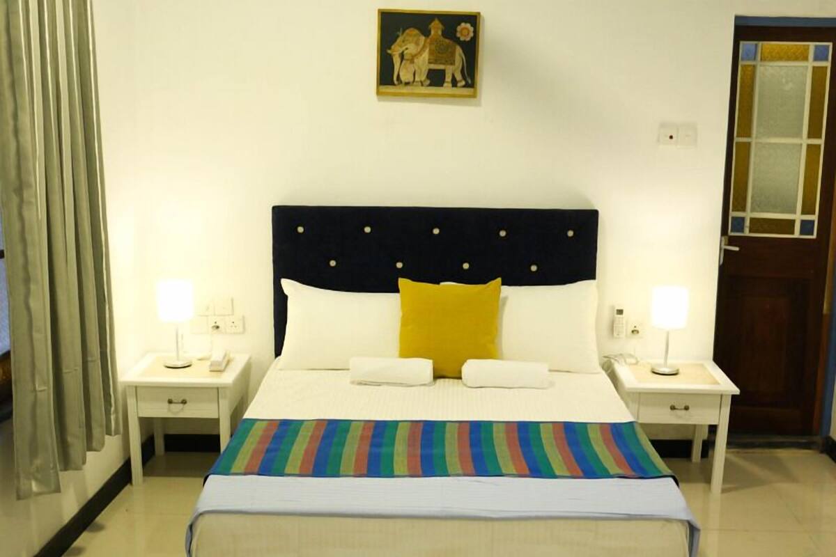 Quarto de hotel com cama, duas mesas de cabeceira, um criado-mudo, um quadro pendurado na parede e uma porta.