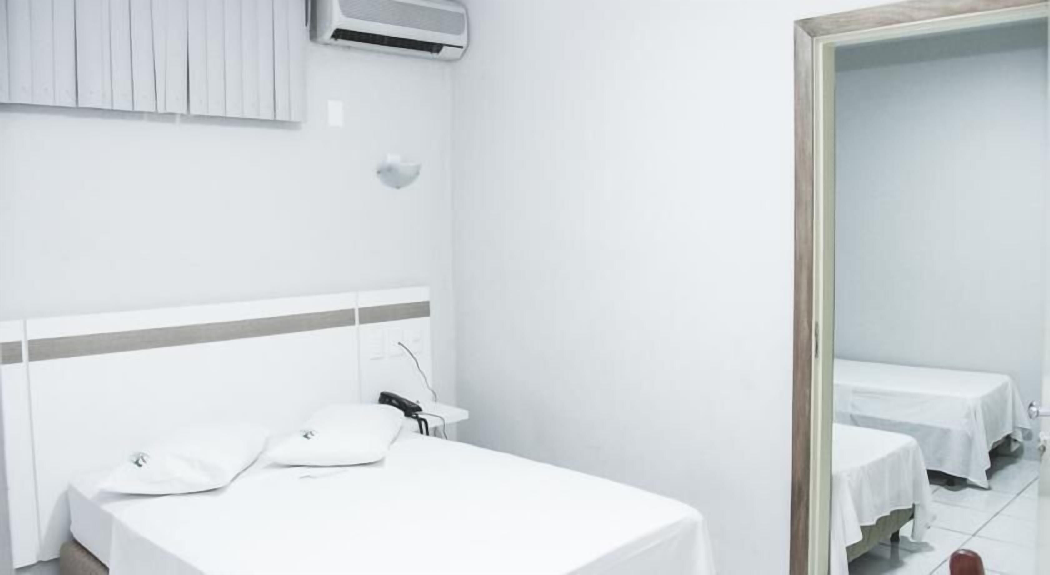 Quarto de hotel com uma cama de solteiro, uma mesa de cabeceira, um espelho e uma janela com persianas.