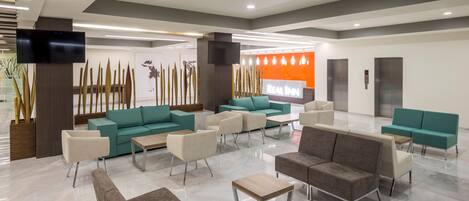 Um lobby moderno de hotel com assentos em tons de verde-água e bege, uma televisão de tela plana e um balcão de recepção.