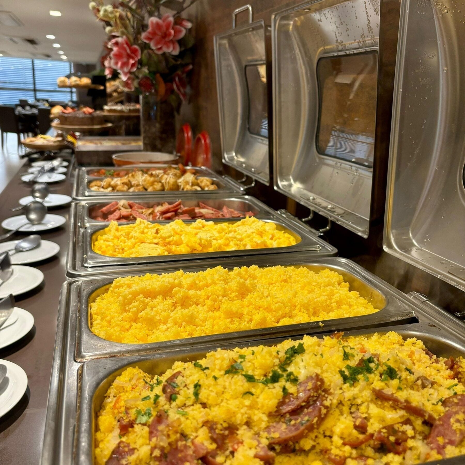 Buffet com diversas opções de comida, incluindo ovos mexidos, pão de milho e carnes.