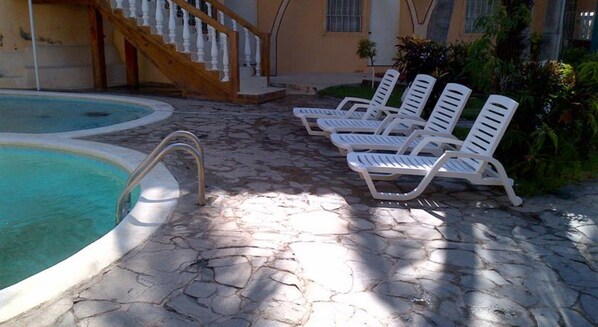 Uma área de piscina com deck de pedra, escada de madeira e espreguiçadeiras brancas.